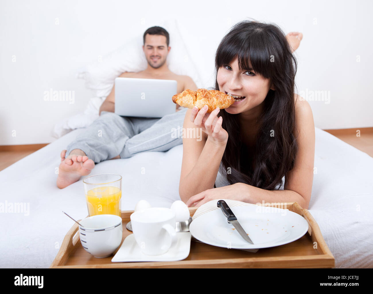 Young woman eating breakfast in bed while man using laptop Stock Photo - Alamy