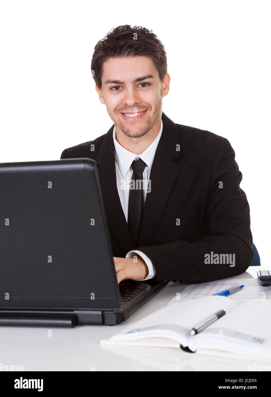 Smiling handsome young businessman seated at his desk behind his laptop ...