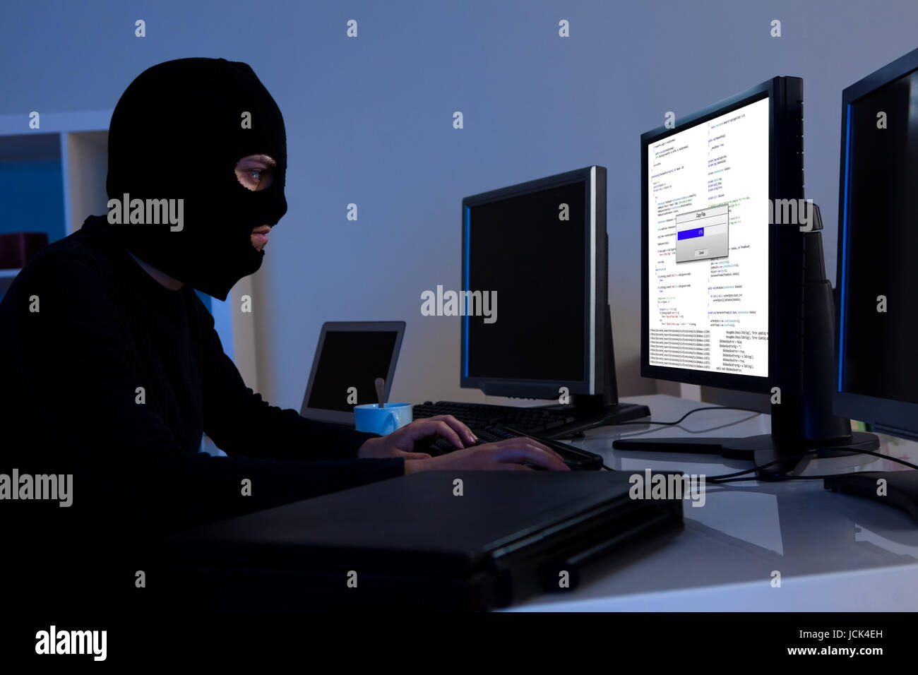 Masked hacker wearing a balaclava sitting at a desk downloading private ...