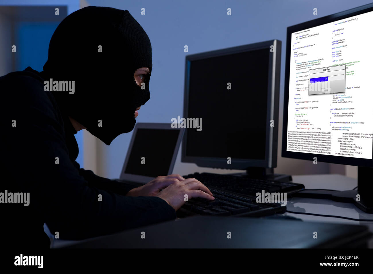 Masked hacker wearing a balaclava sitting at a desk downloading private ...