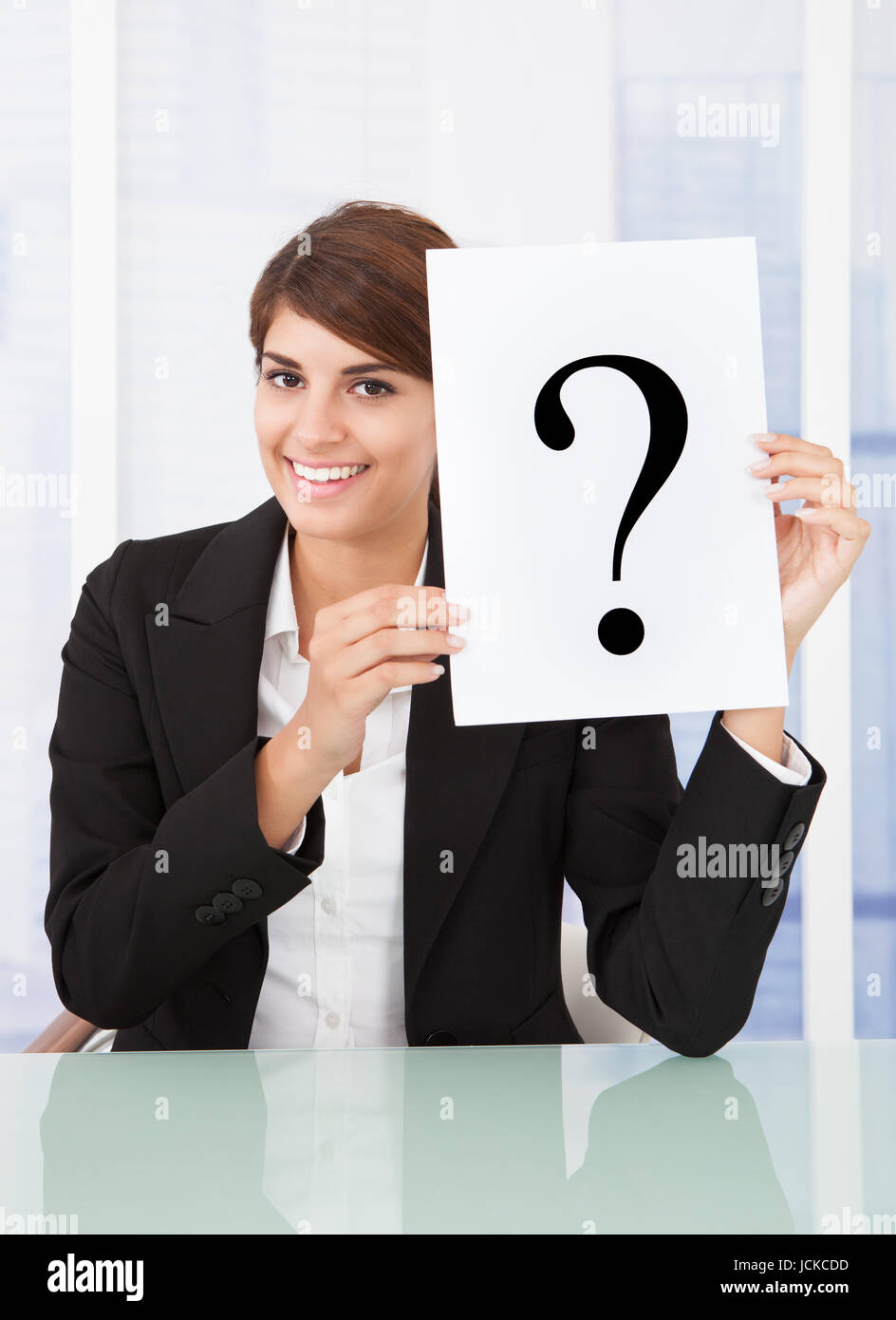 Businesswoman holding paper with question mark in front of face at desk ...