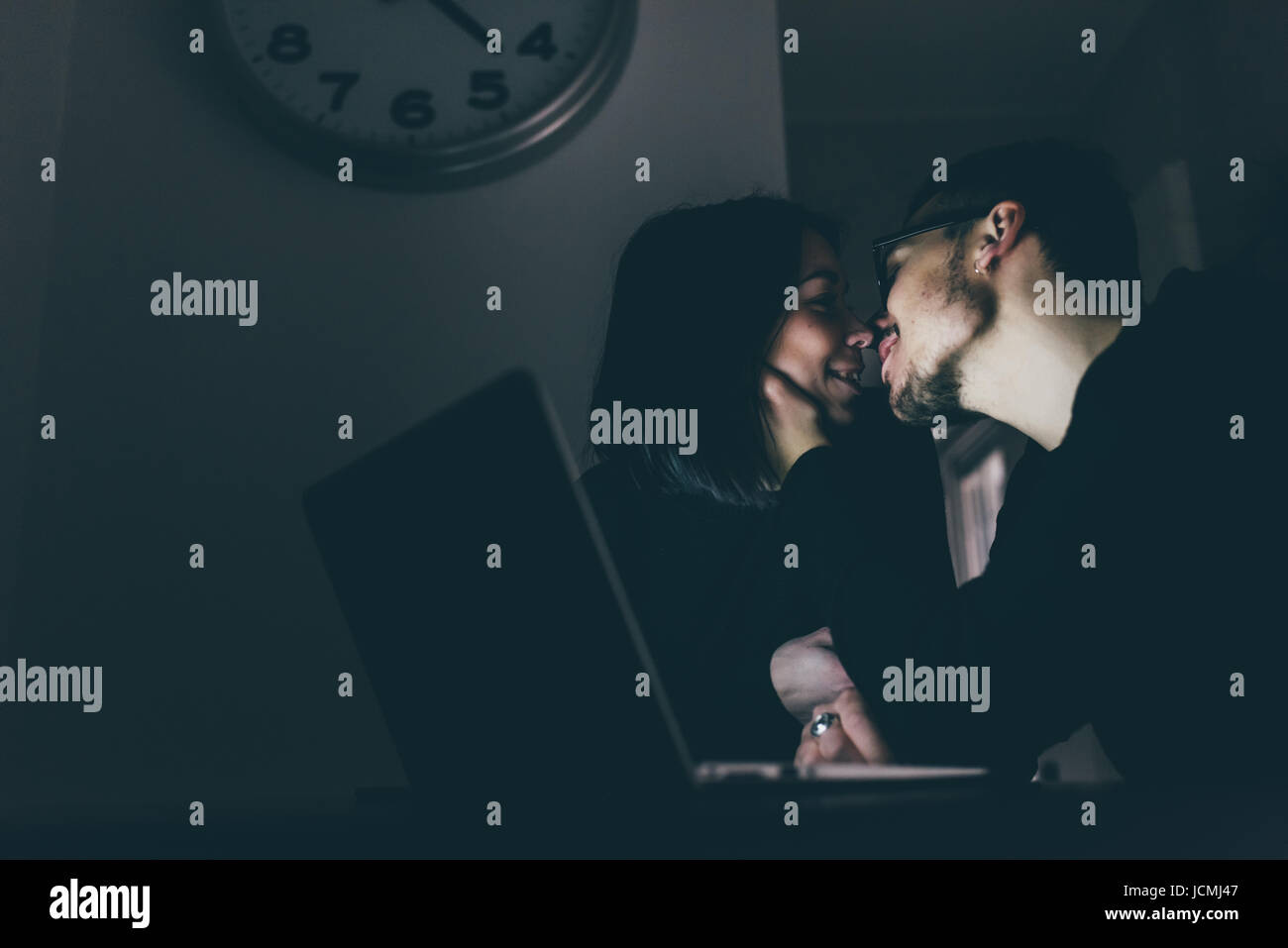 Young couple kissing using computer illuminated by screen light smiling ...