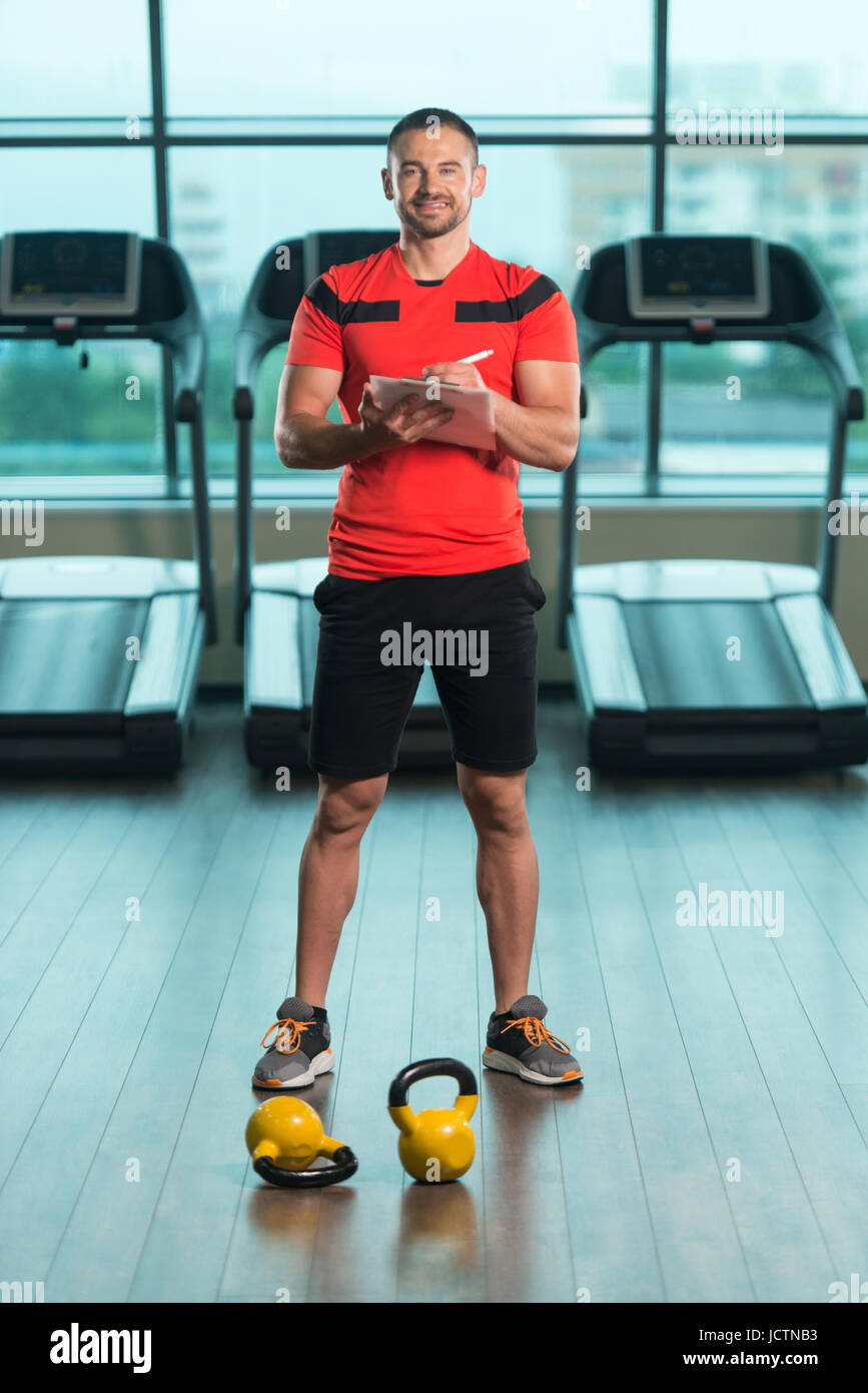 Handsome Personal Trainer With A Clipboard In The Gym And Flexing ...