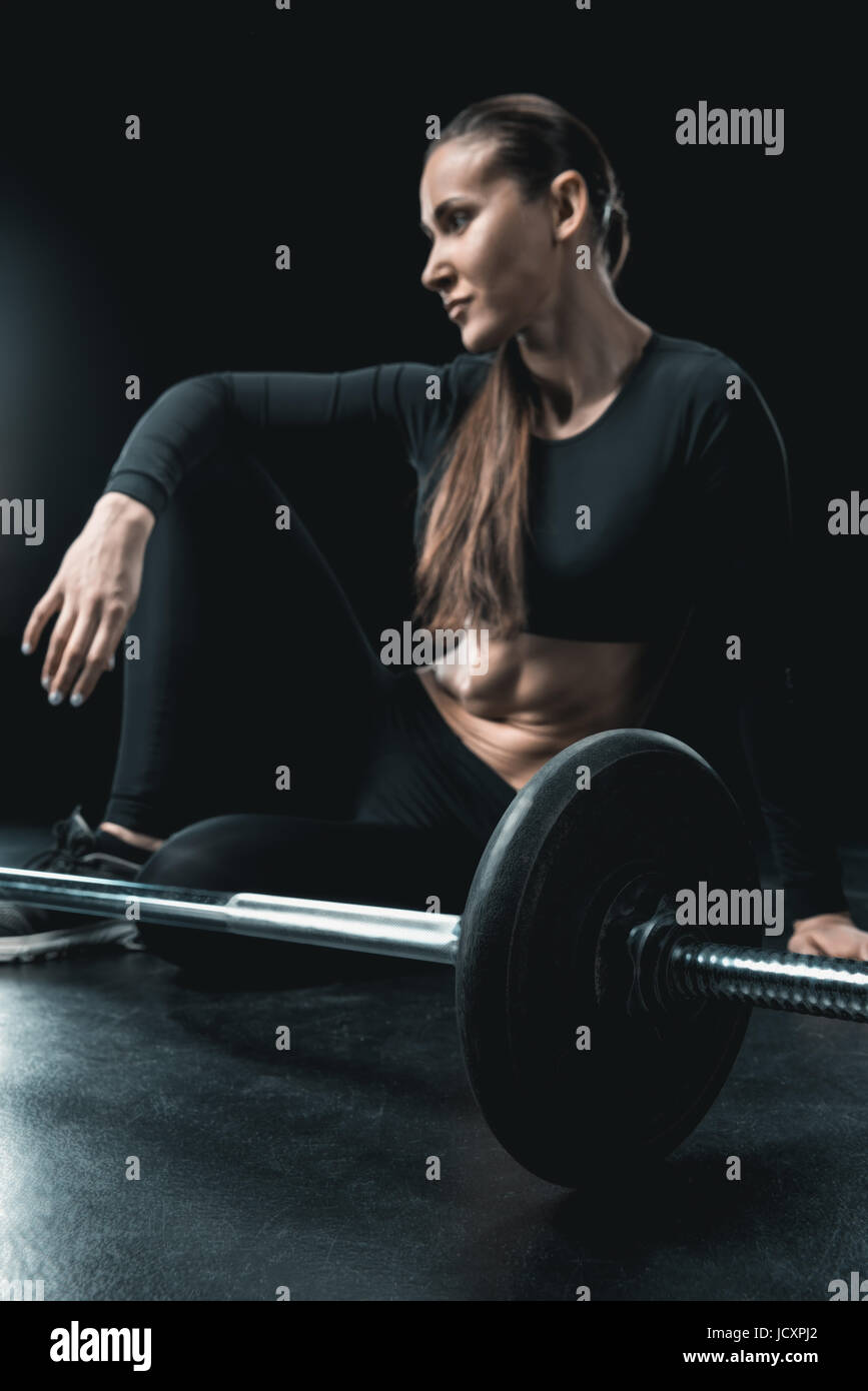 young fitness woman sitting at barbell and resting isolated on black ...