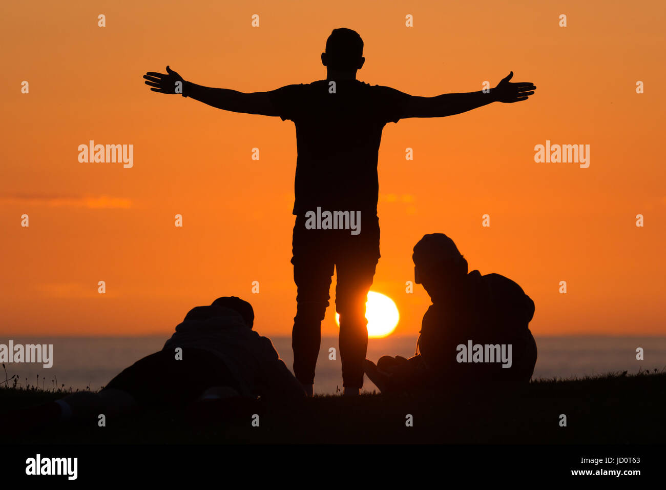 A silhouetted man standing with his arms out stretched while the sun sets over the west coast ...