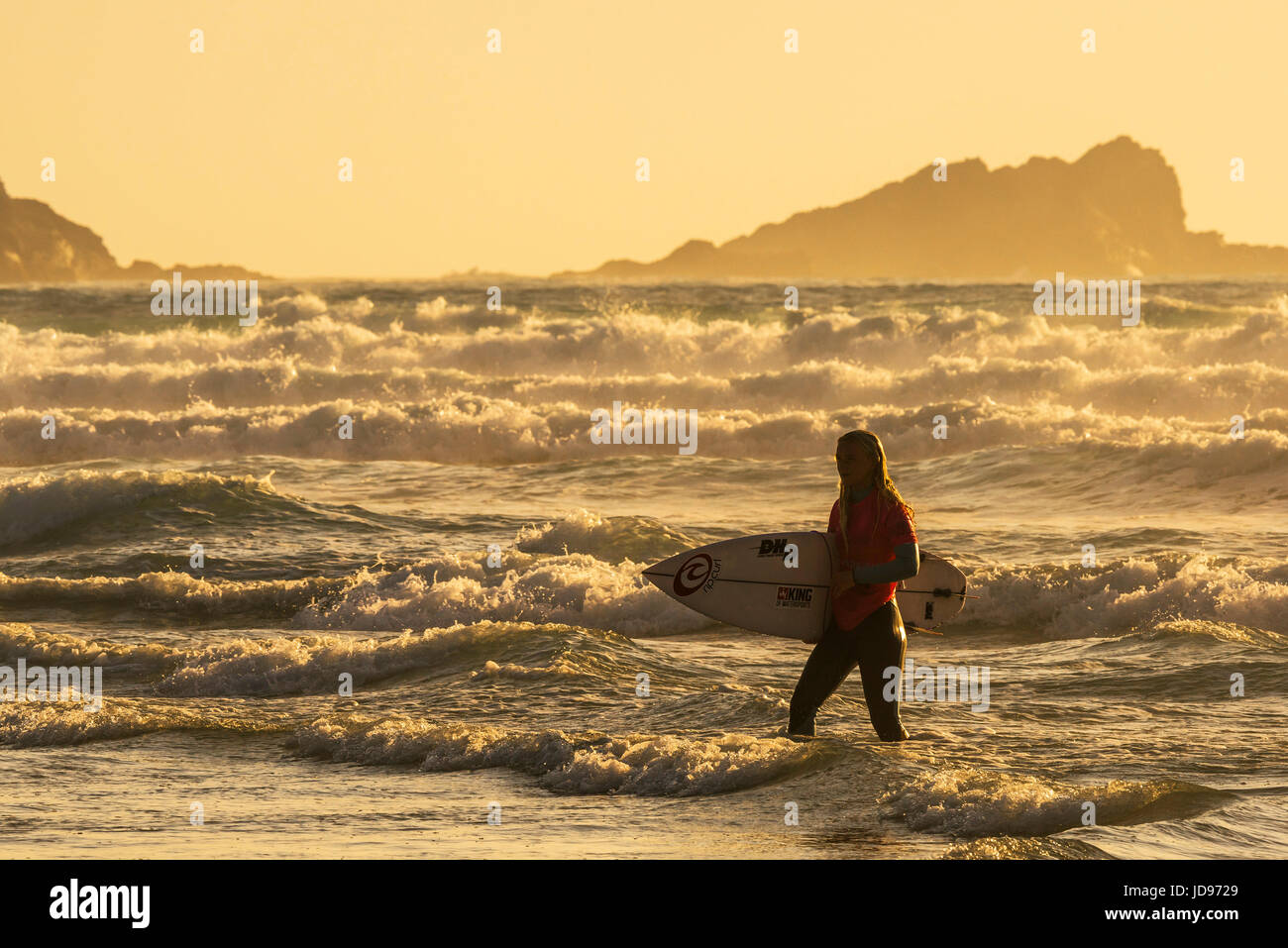 A female surfer walks from the sea at sunset. Fistral Beach, Newquay ...