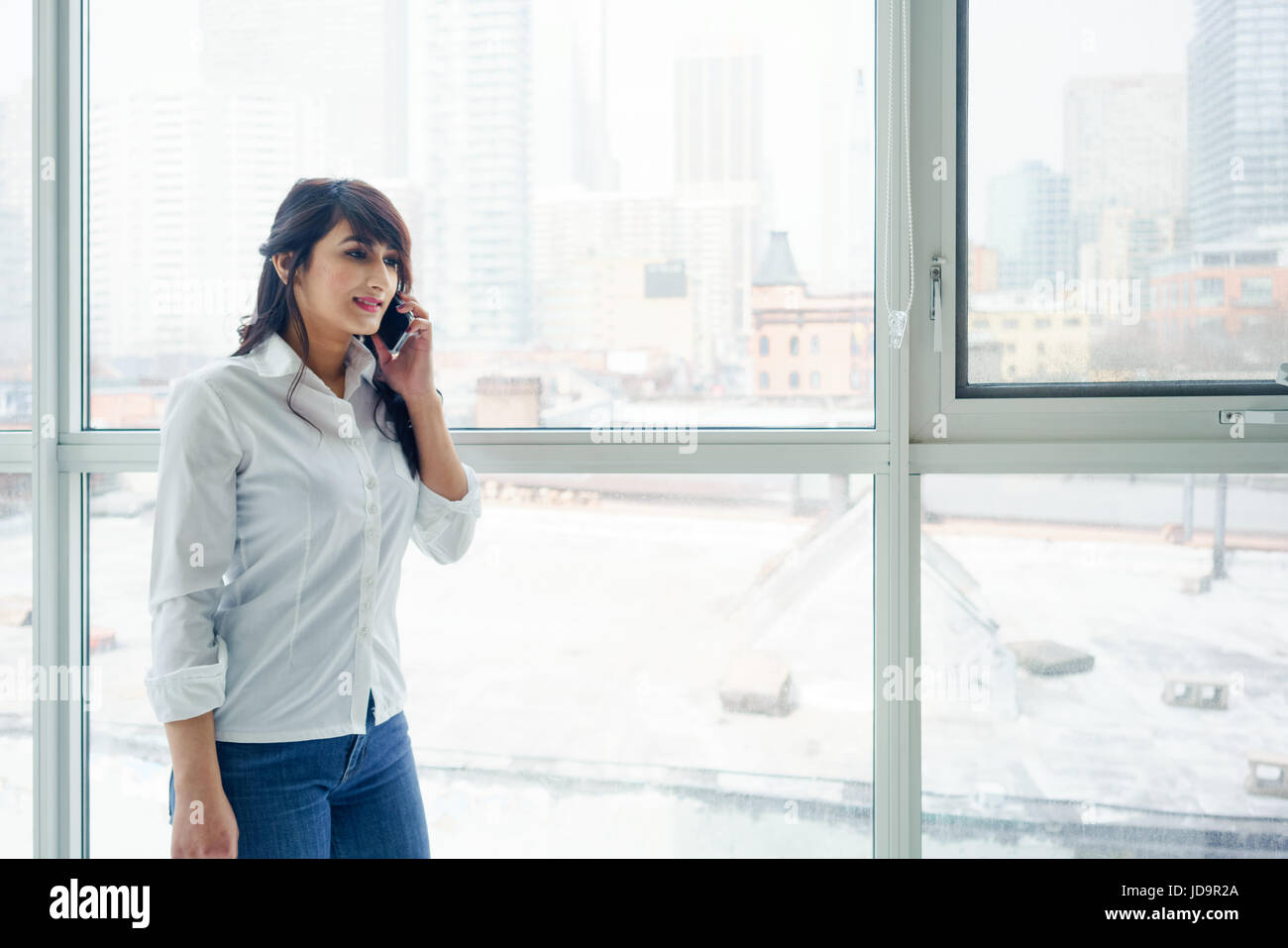 Young woman indoors, standing next to window using mobile phone. young ...