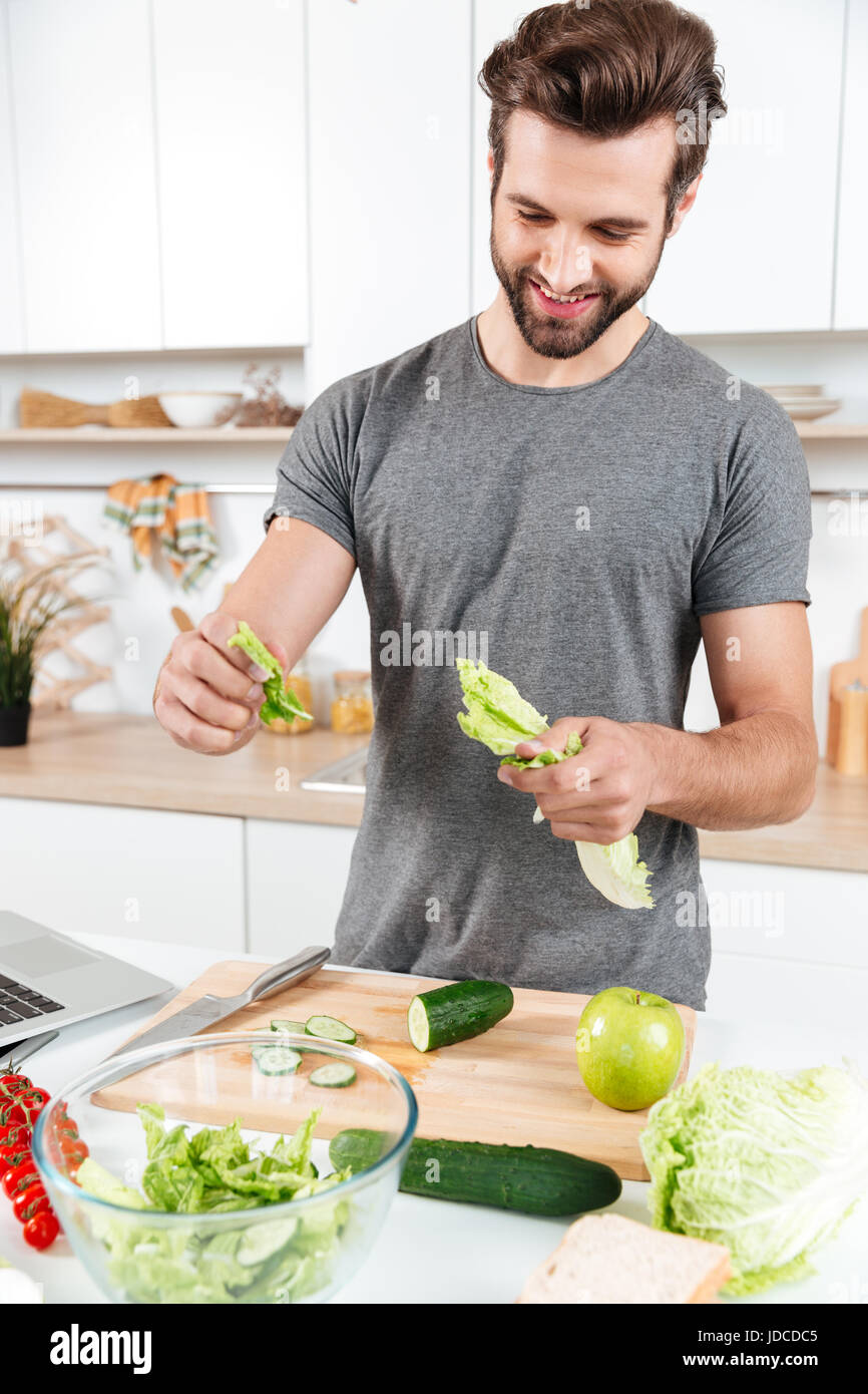 Happy handsome guy cooking salad with laptop at his modern kitchen at ...