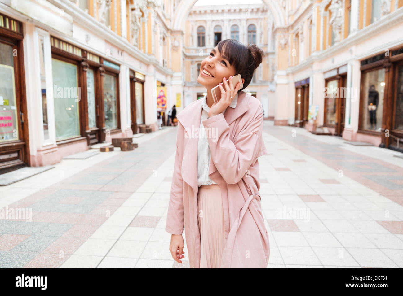 Cheerful cute girl in pink coat talking on mobile phone while walking ...