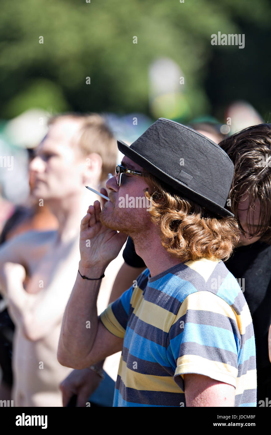 A man with ginger hair and wearing a hat smoking a cigarette at an ...