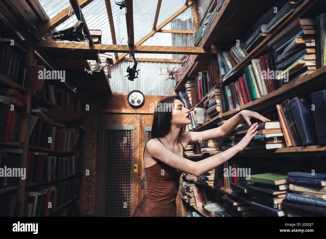 Side view of young brunette woman standing in library and leaning on ...