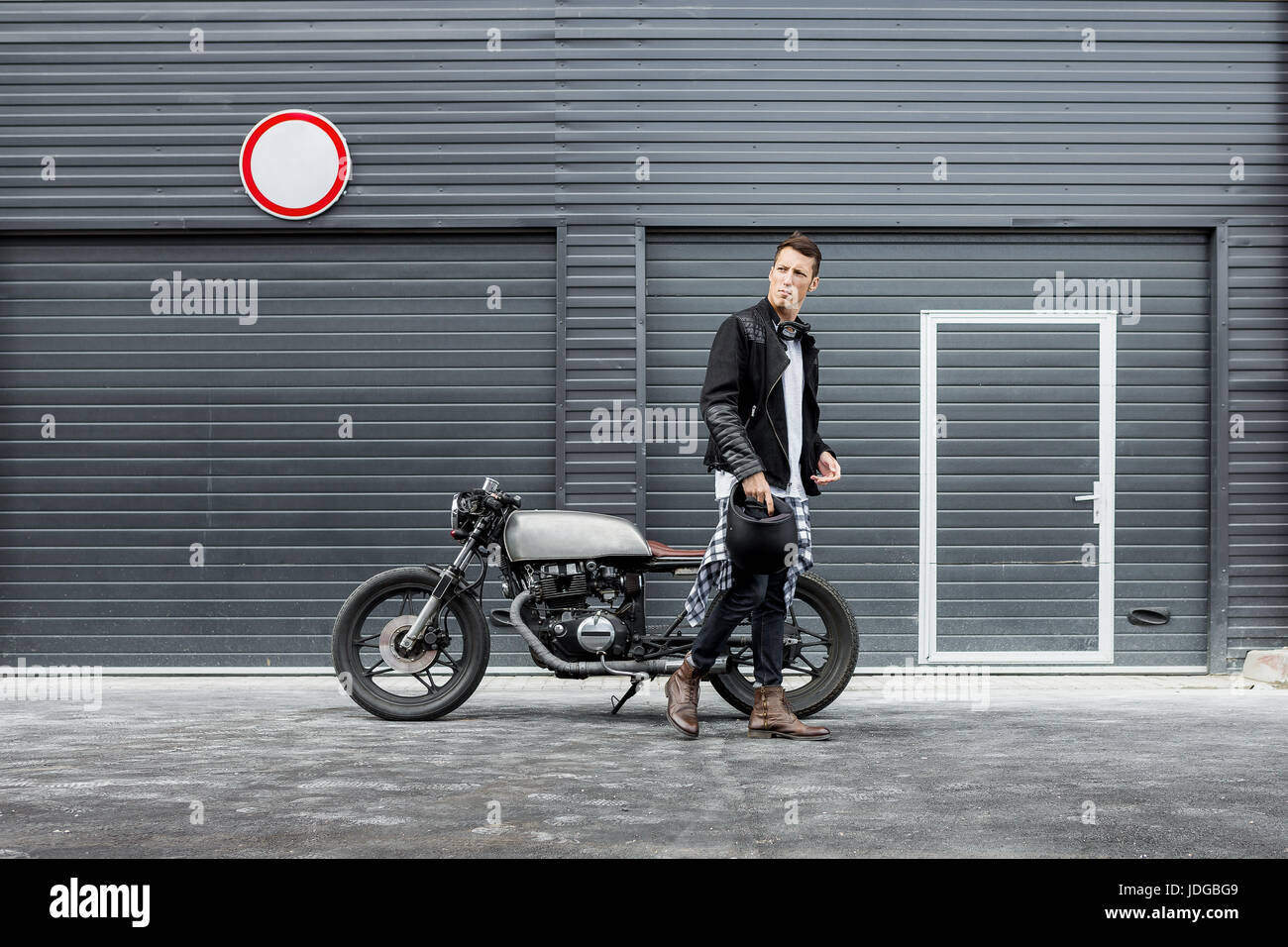 Handsome rider biker guy hold his black helmet near industrial gates ...