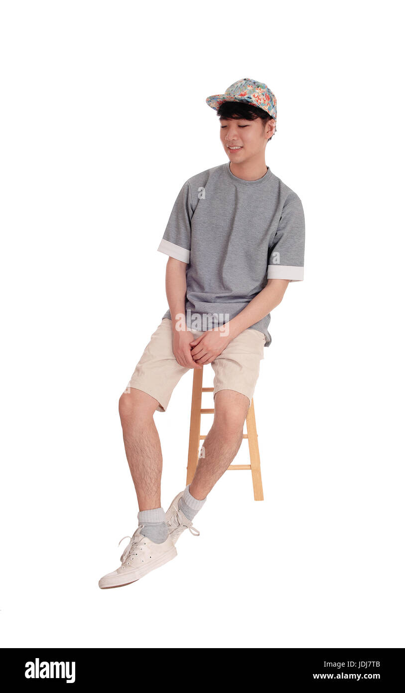 A young Asian teenager sitting on a chair in shorts and a cap, smiling ...