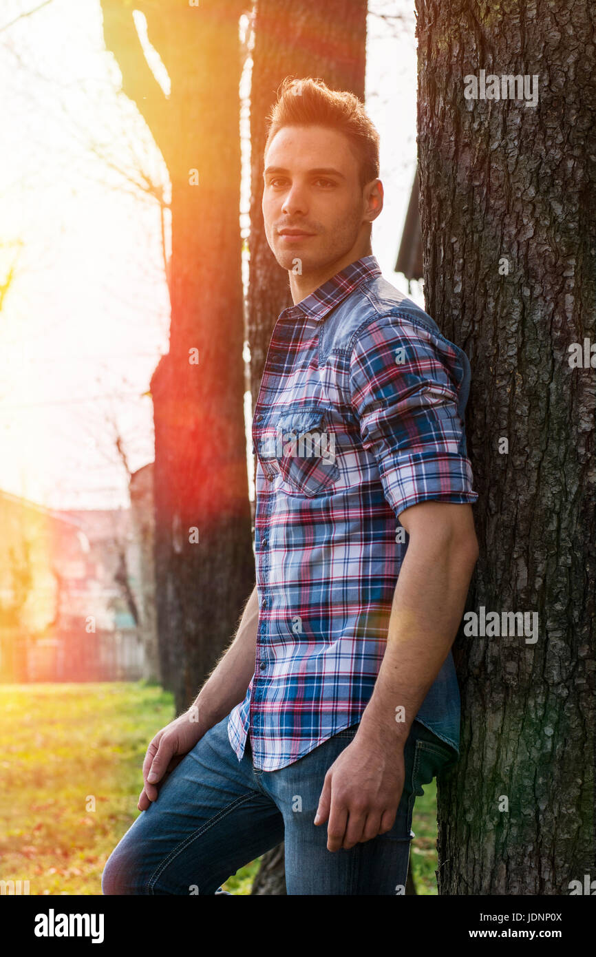 A young man outdoors against a tree, at sunset, with sun behind him ...