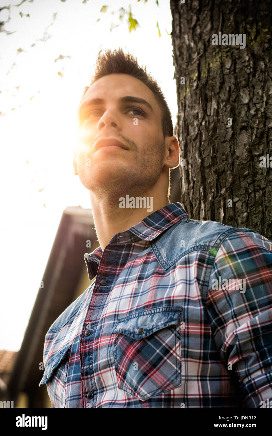 A young man outdoors against a tree, at sunset, with sun behind him ...
