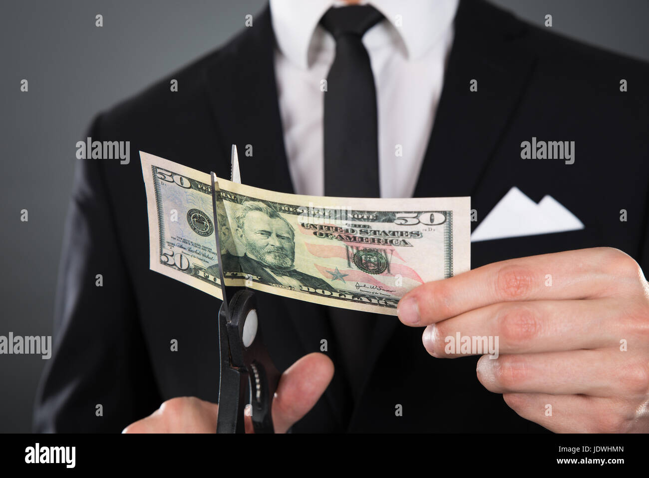 Midsection of businessman cutting dollar bill with scissors over gray ...