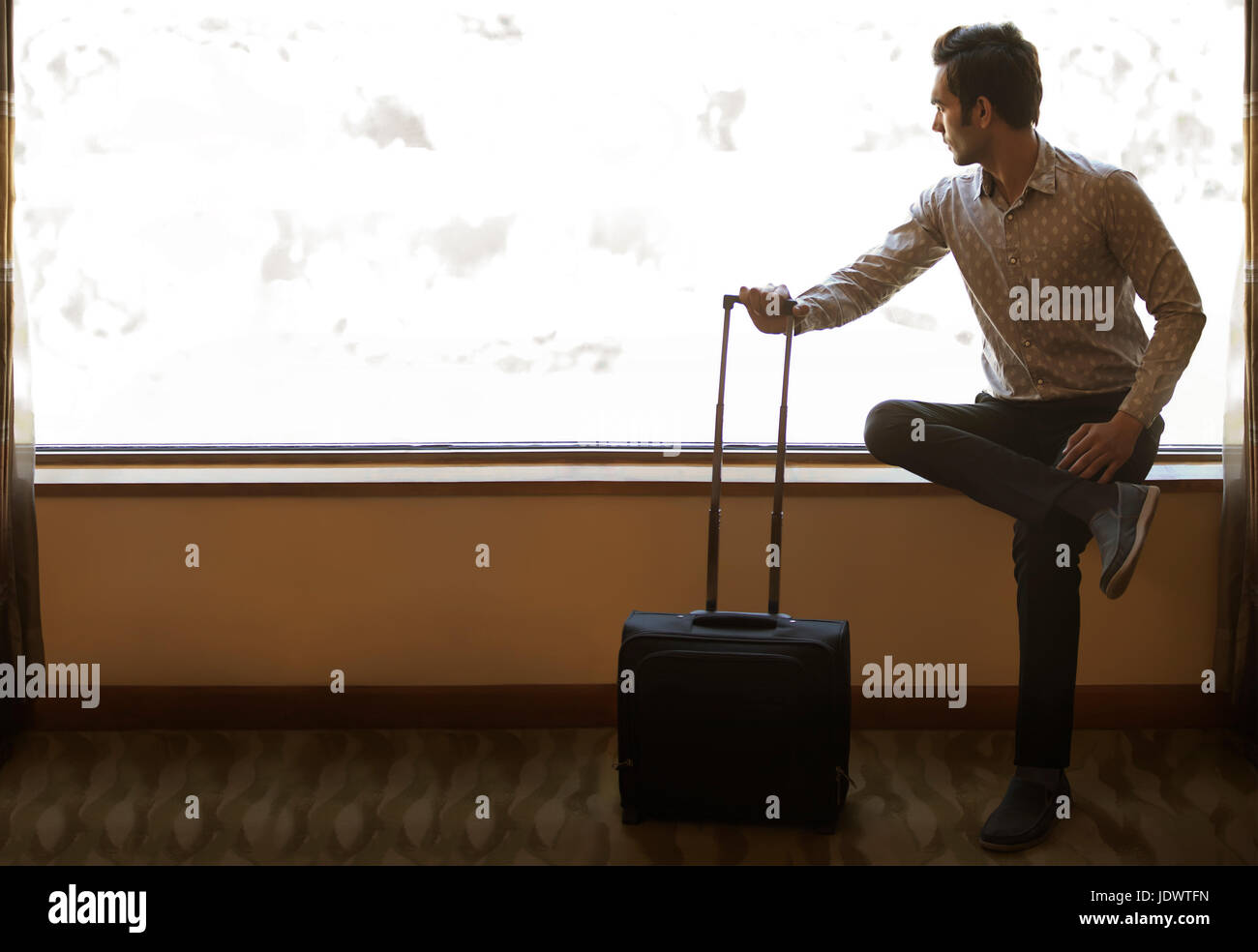 Young man sitting near window holding trolley bag Stock Photo - Alamy