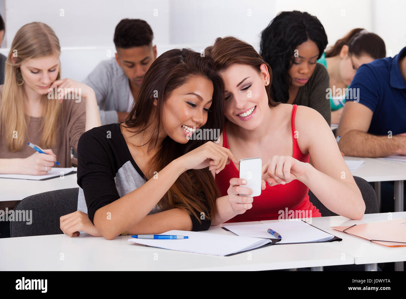 Happy female college students using mobile phone together at desk in ...