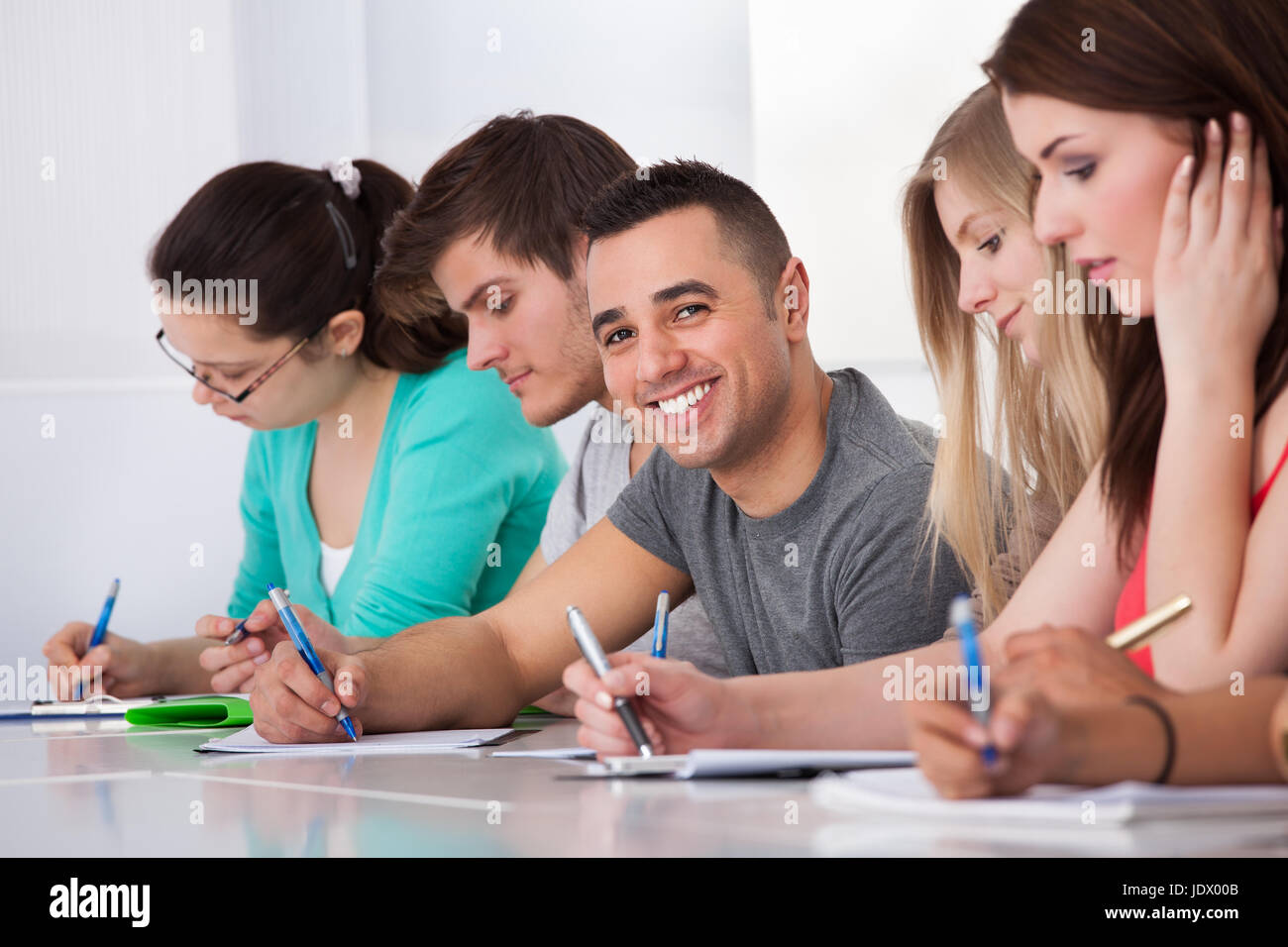 Portrait of handsome university student sitting with classmates writing ...