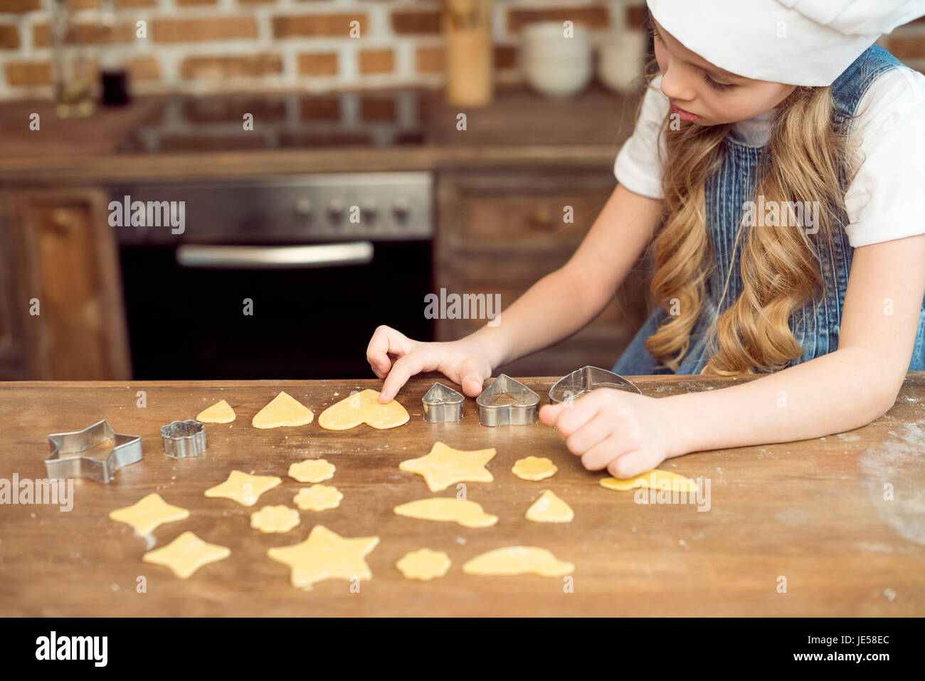 side view of little girl in chef hat making shaped cookies in kitchen ...