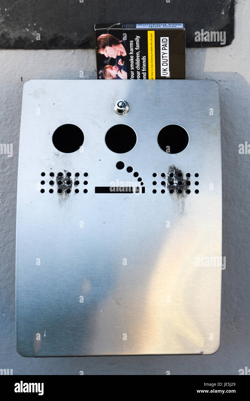 Wall mounted outdoor ashtray showing signs of use. A cigarette carton ...