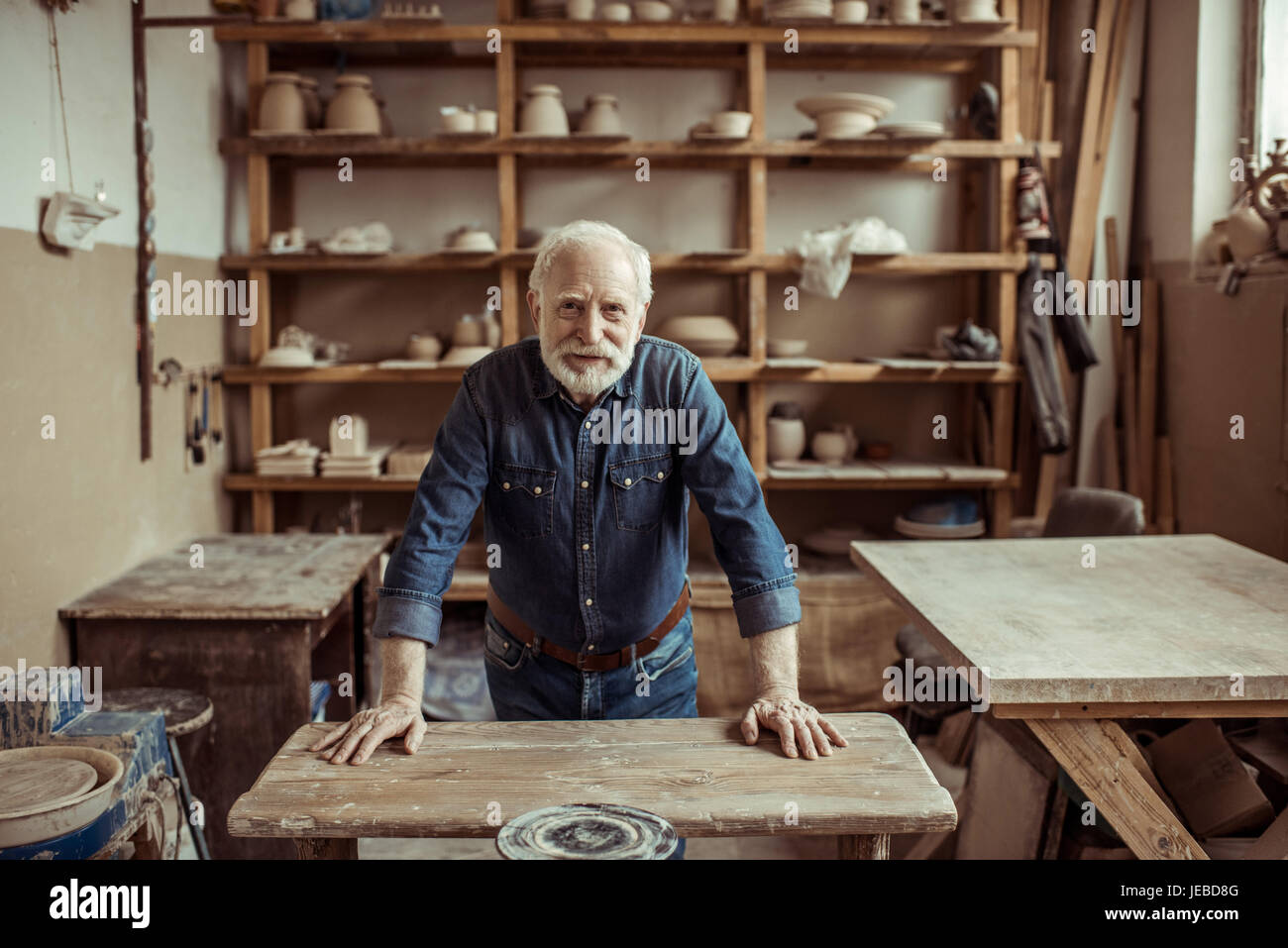 Front view of senior potter standing and leaning on table against ...