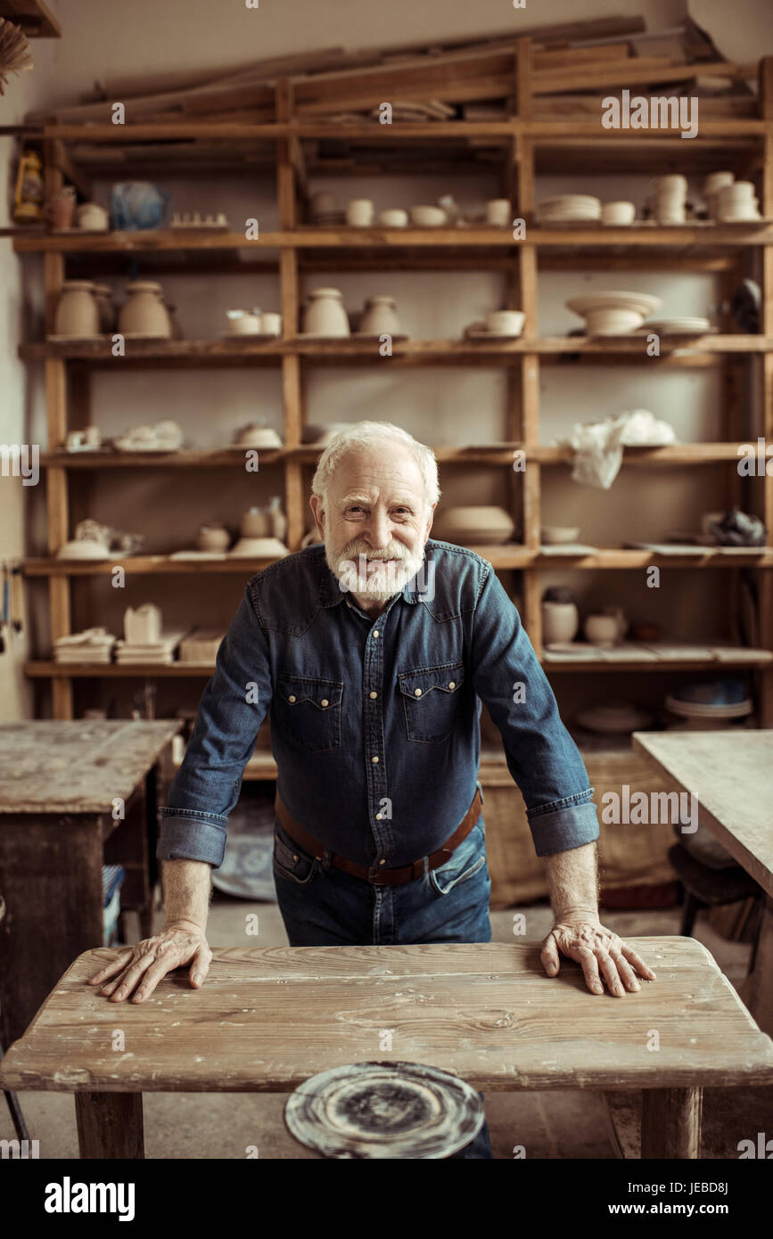 Front view of senior potter standing and leaning on table against ...