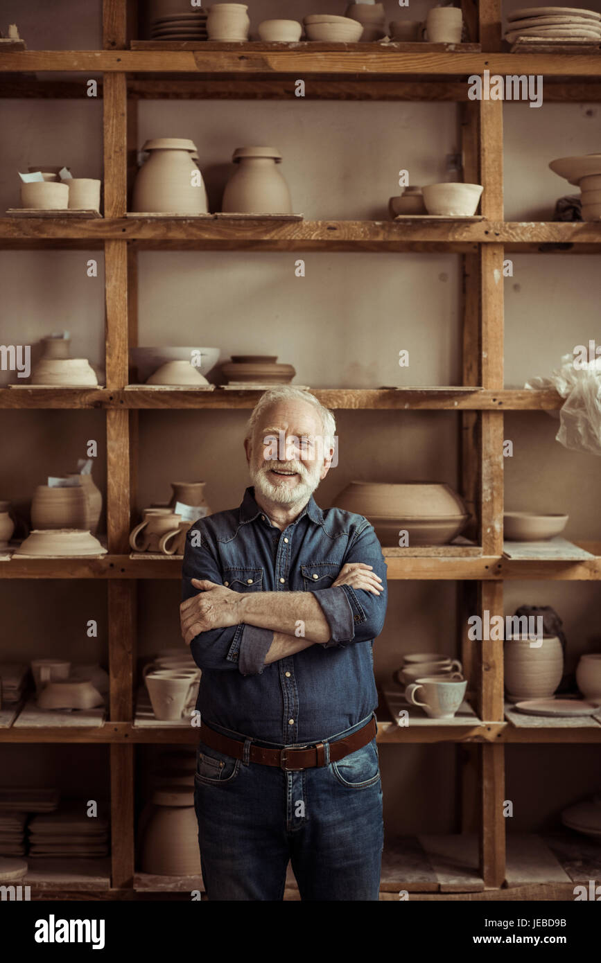 Front view of senior potter standing against shelves with pottery goods ...