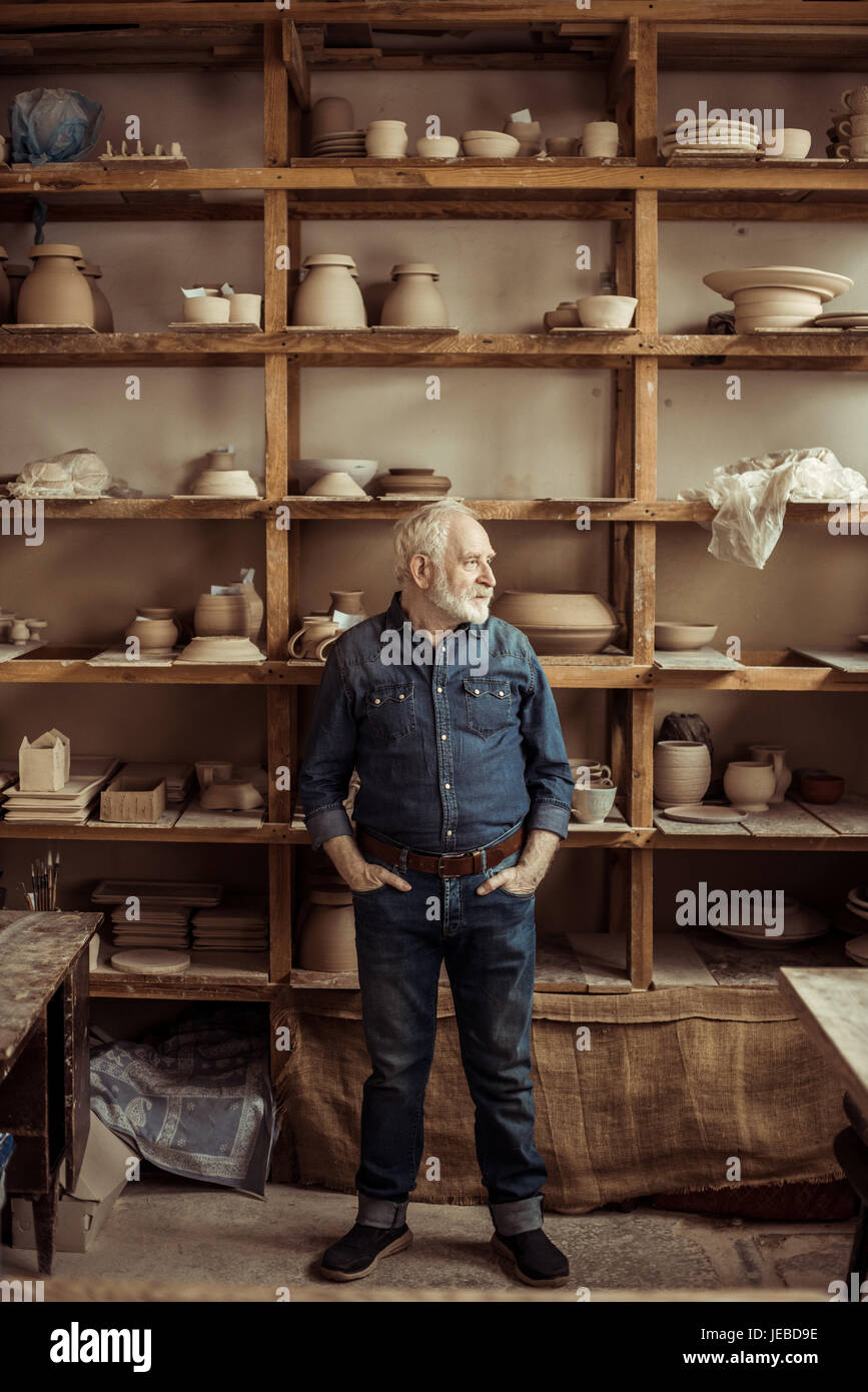 Front view of senior potter standing against shelves with pottery goods ...