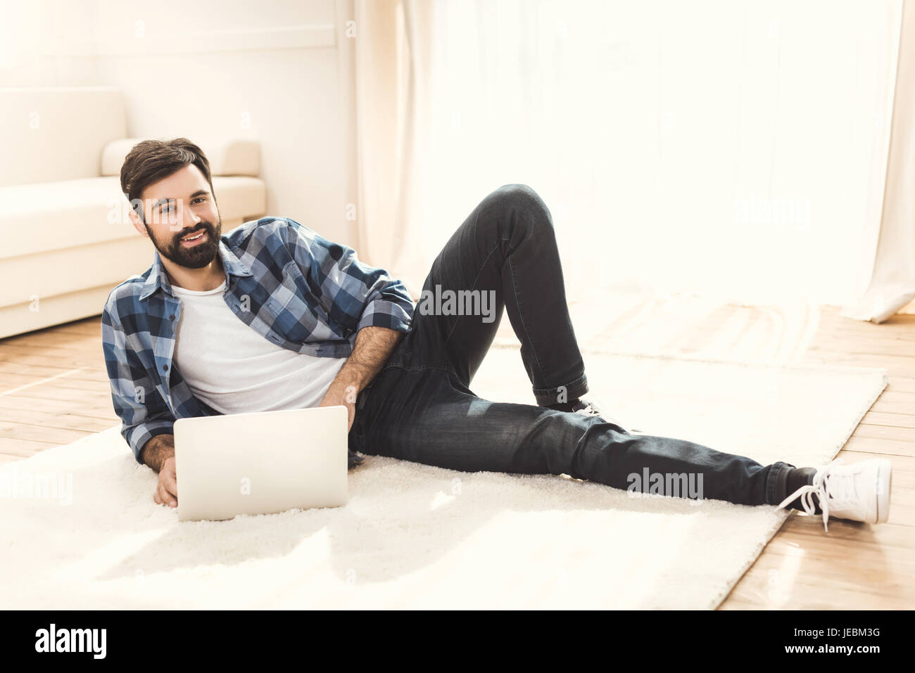 Young bearded man lying on carpet and using laptop computer at home ...