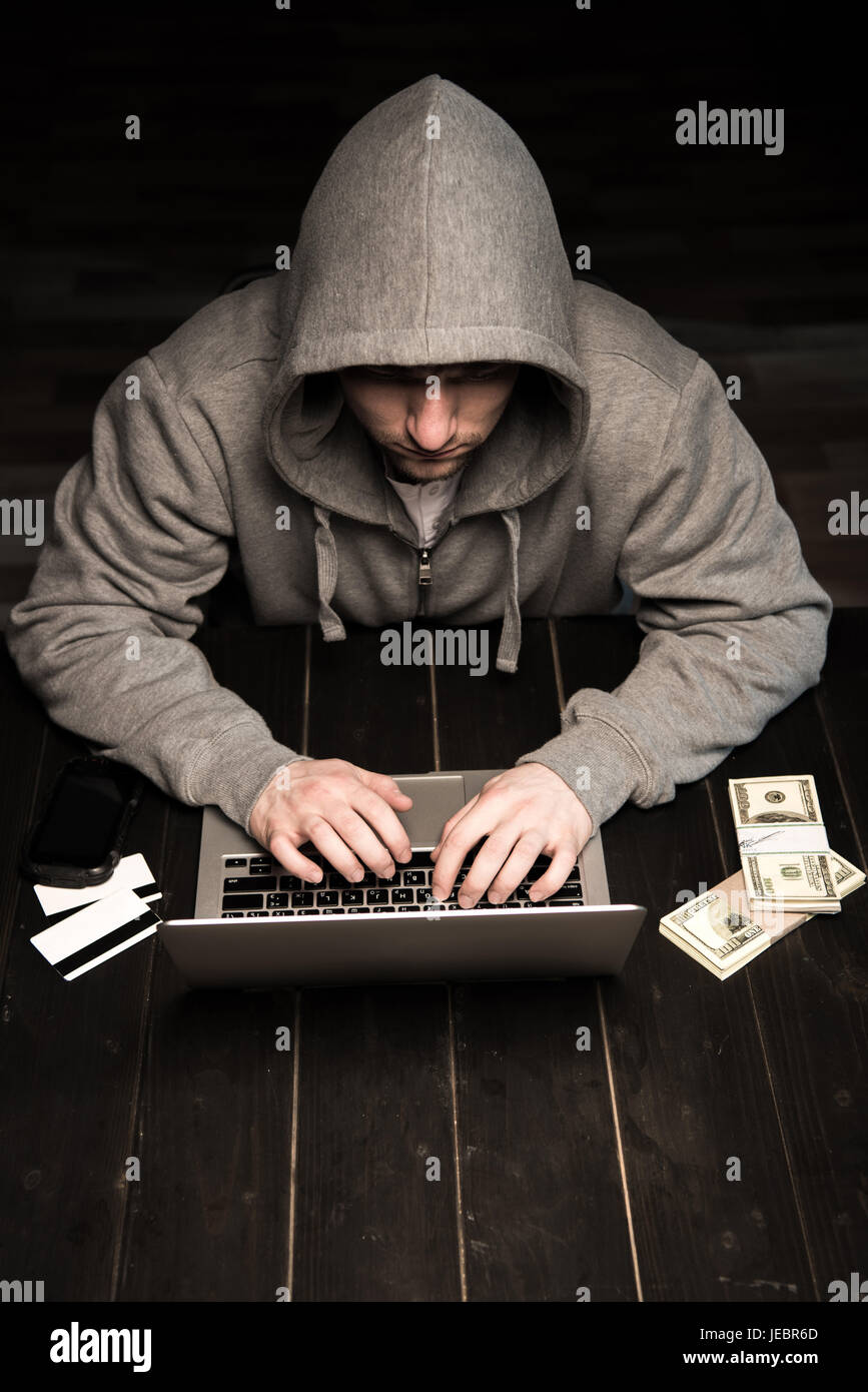 Young hacker in hoodie sitting at table and using laptop Stock Photo ...
