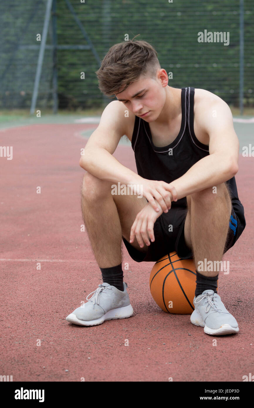 Teenage boy sitting on a basketball on a court looking sad Stock Photo ...