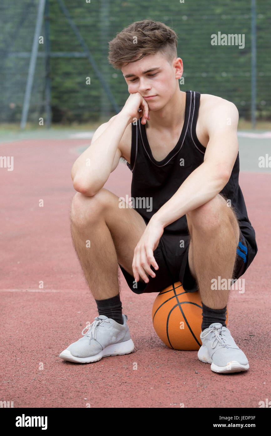 Teenage boy sitting on a basketball on a court looking sad Stock Photo ...