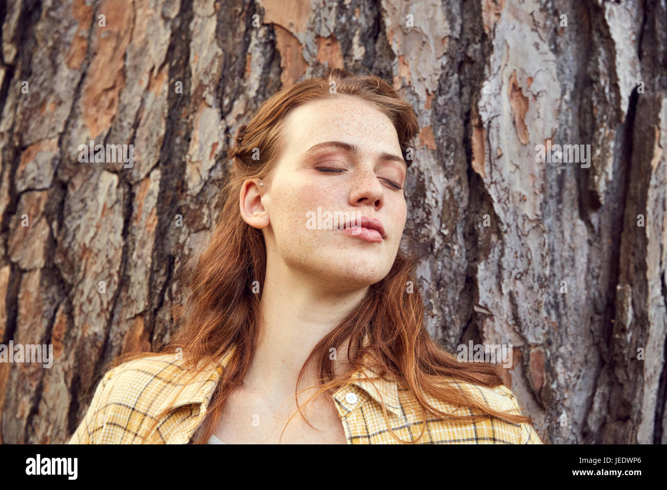 Portrait Of Redheaded Young Woman Leaning Against Tree Trunk With Eyes