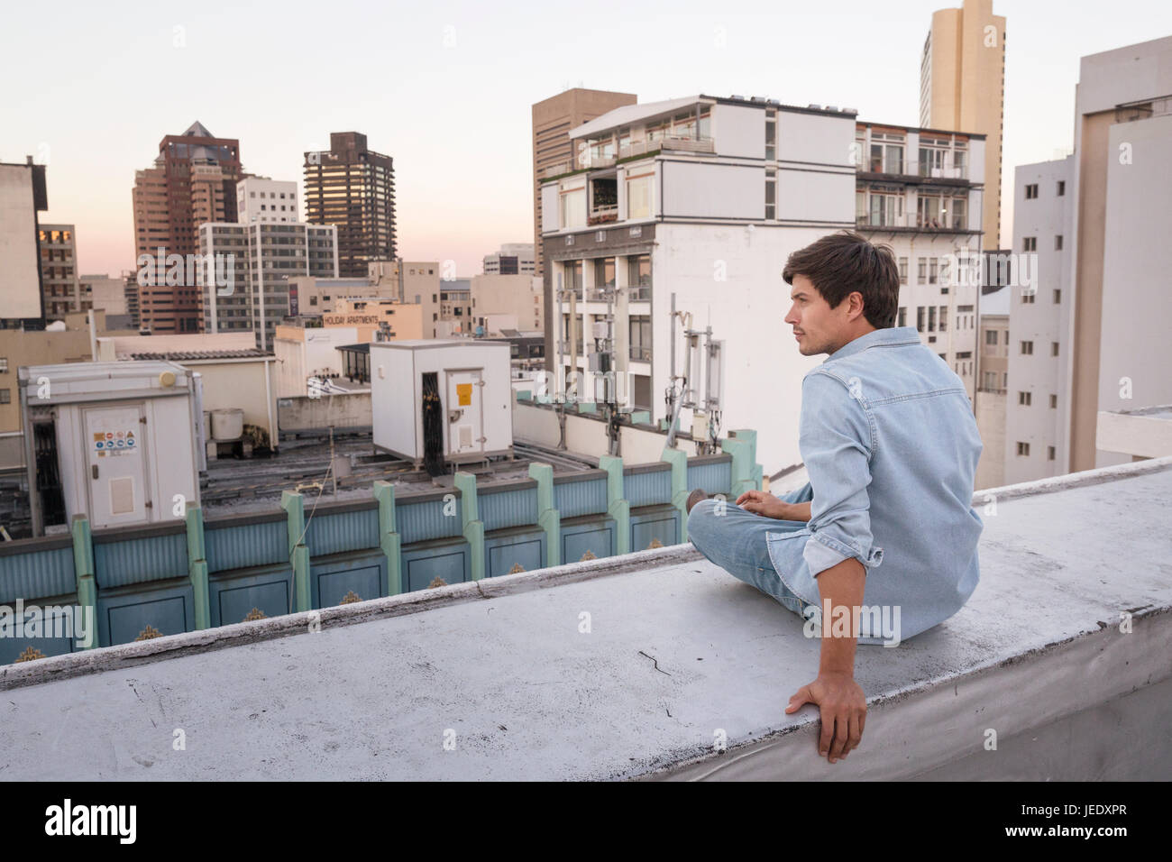 Young man sitting on balustrade of a rooftop terrace, looking at view ...