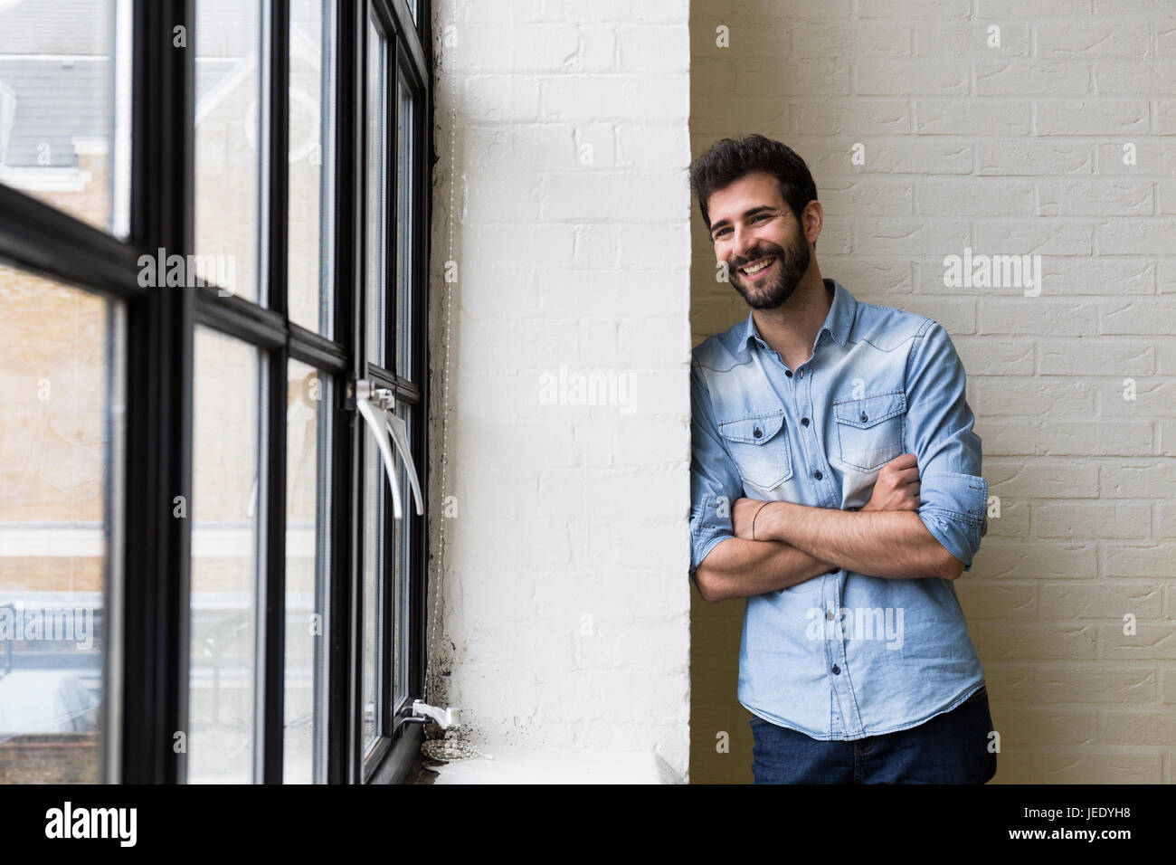 Smiling man leaning against a wall in his loft looking through window ...