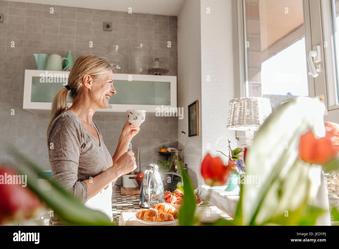 Happy woman at home in kitchen looking out of window Stock Photo - Alamy