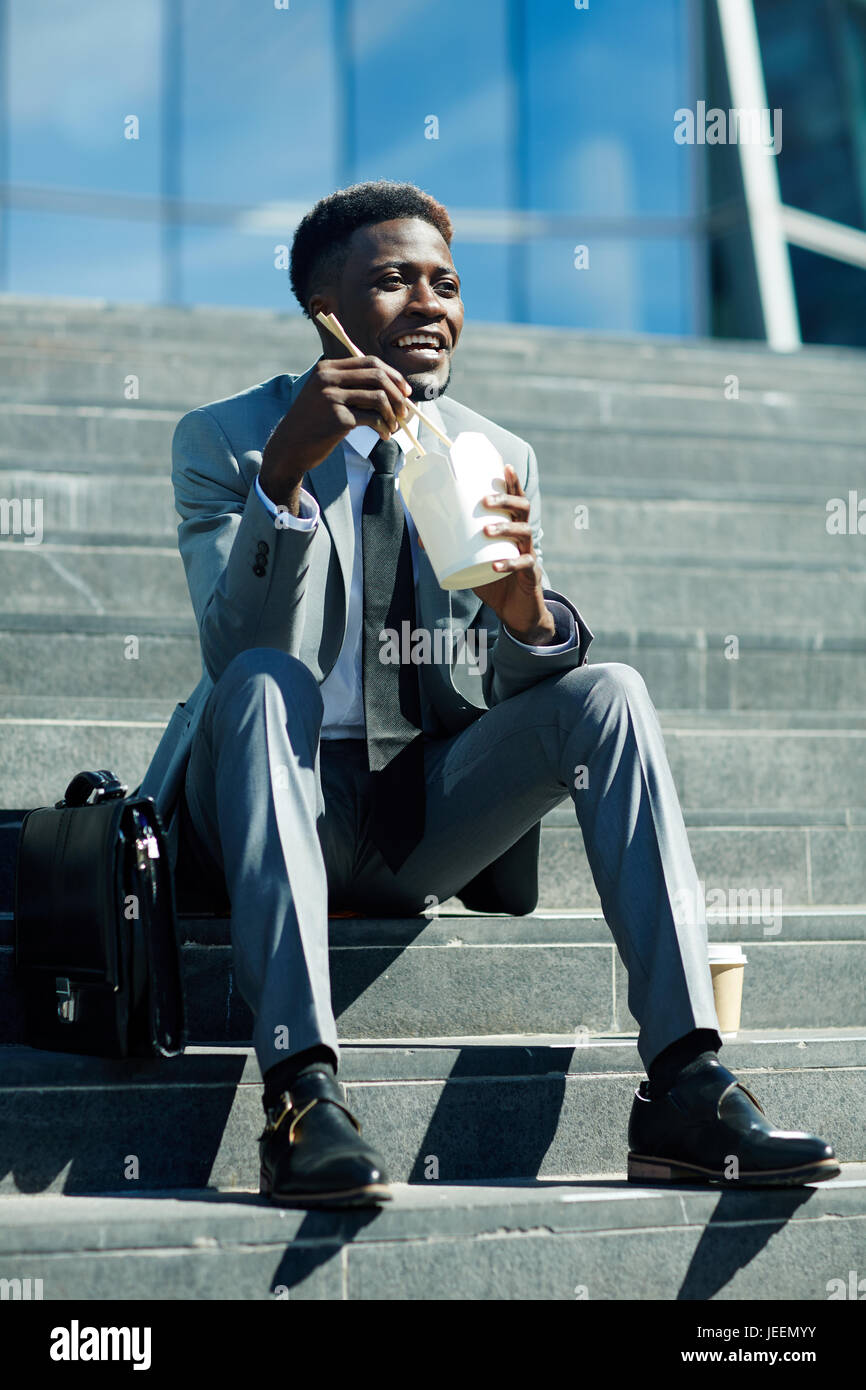 Happy businessman with chopsticks eating food brom box on staircase ...