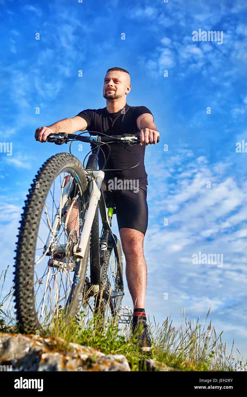 Bottom view of young cyclist standing with bike on rocks against blue ...