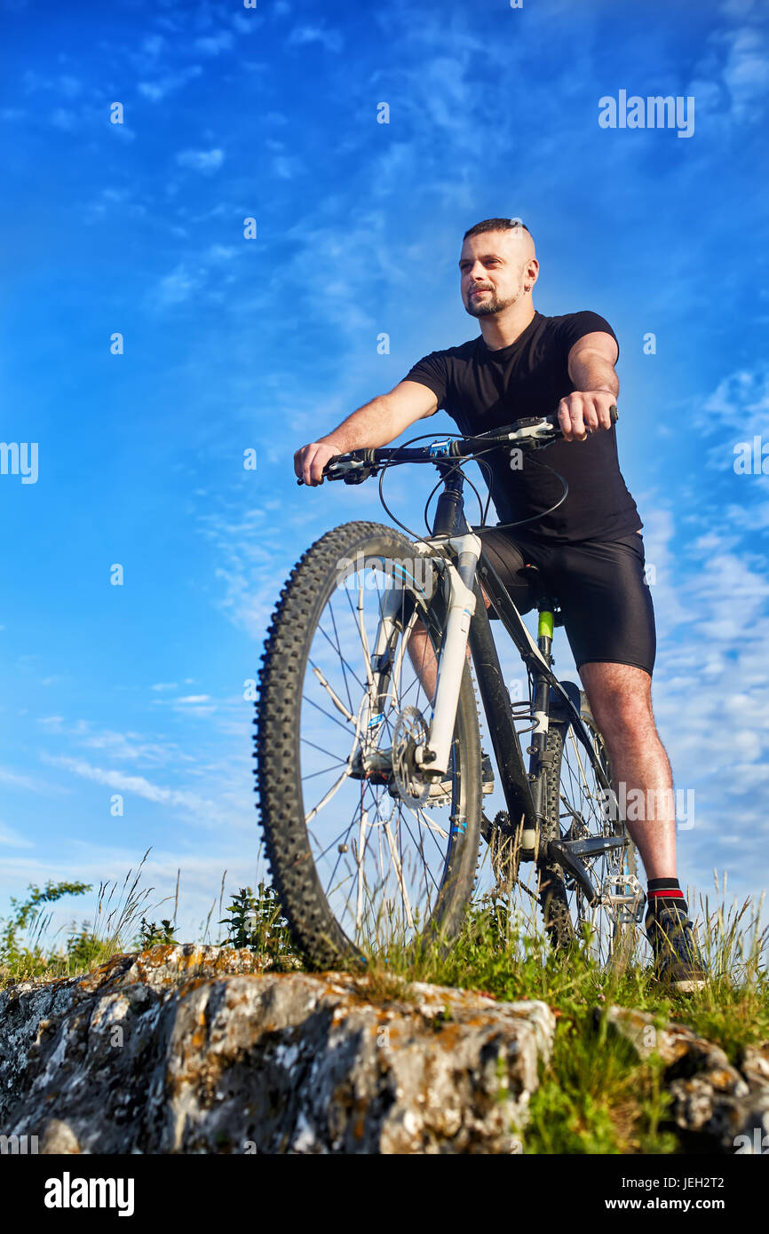 Bottom view of young cyclist standing with bike on rocks against blue ...