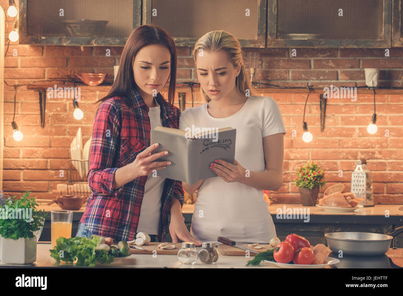 Young female friends cooking meal together at home reading receipt book ...