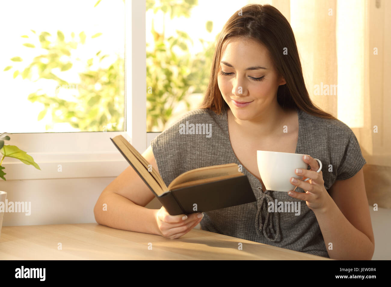 Relaxed woman reading a paper book and holding a cup of coffee sitting ...