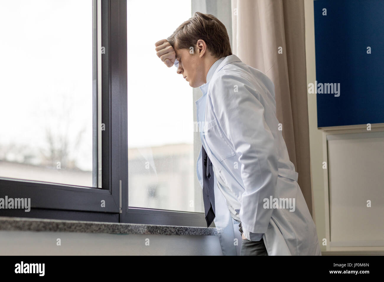 side view of young upset doctor standing and thinking at window in ...