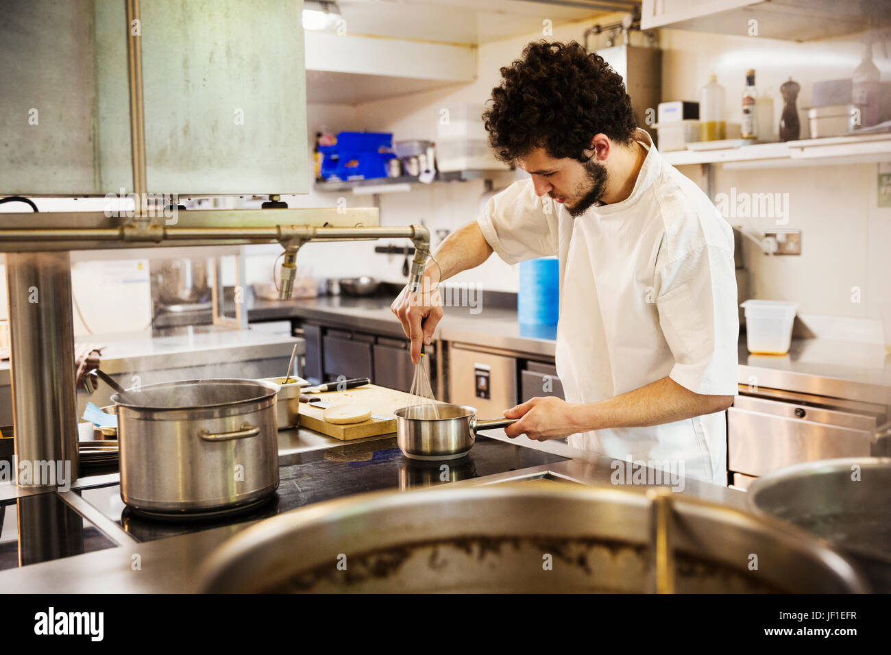 Chef standing in a restaurant kitchen at a stove, holding a whisk ...
