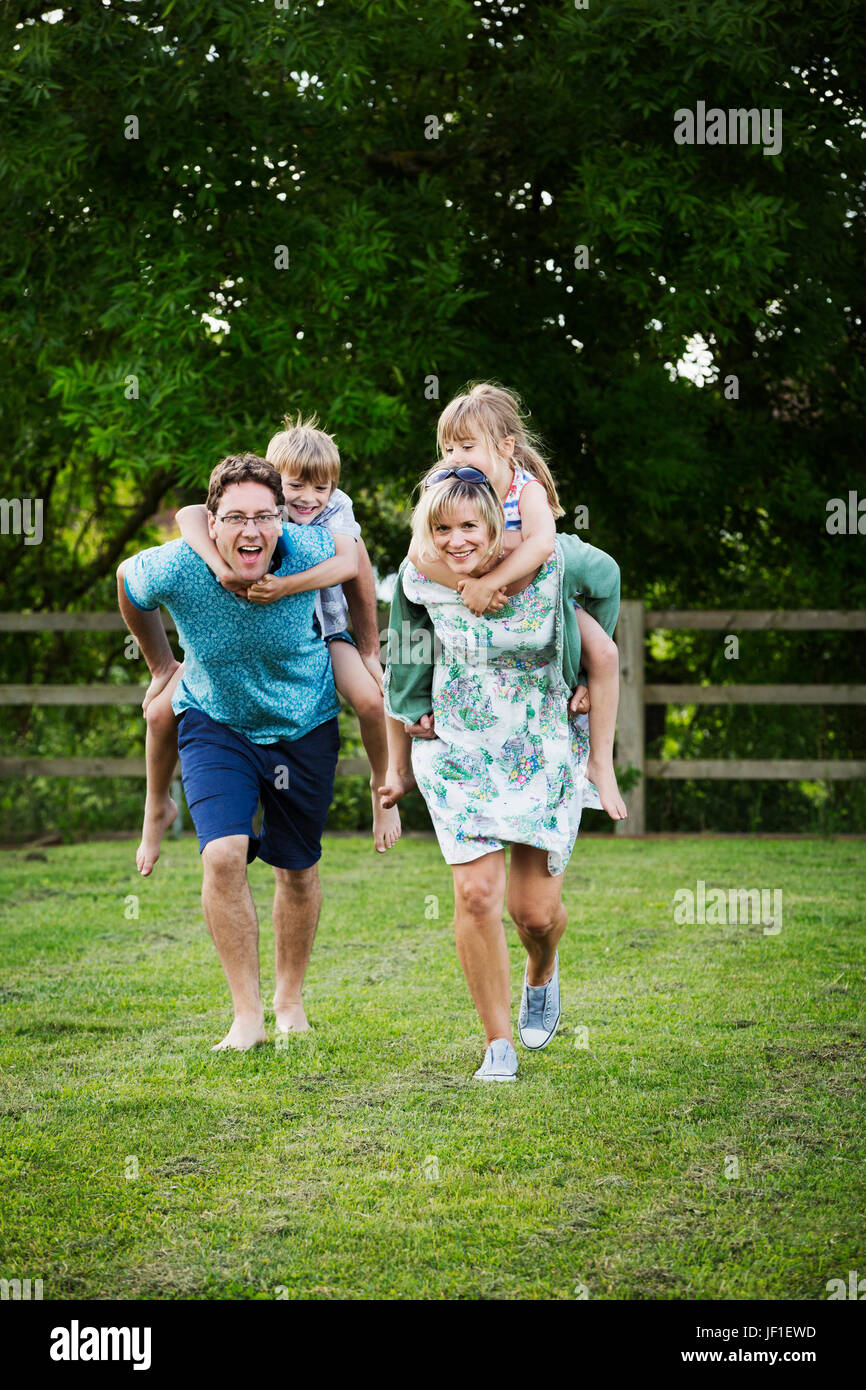 Man and woman running across a lawn, carrying boy and girl piggyback, a ...