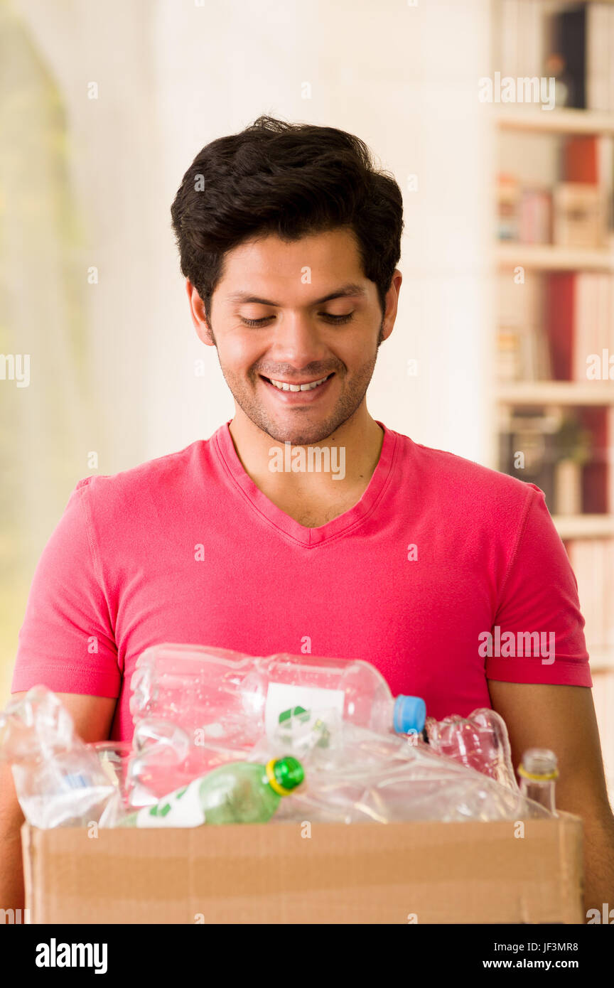 Smiling young man carrying a cardboard box full of plastic, wearing a ...