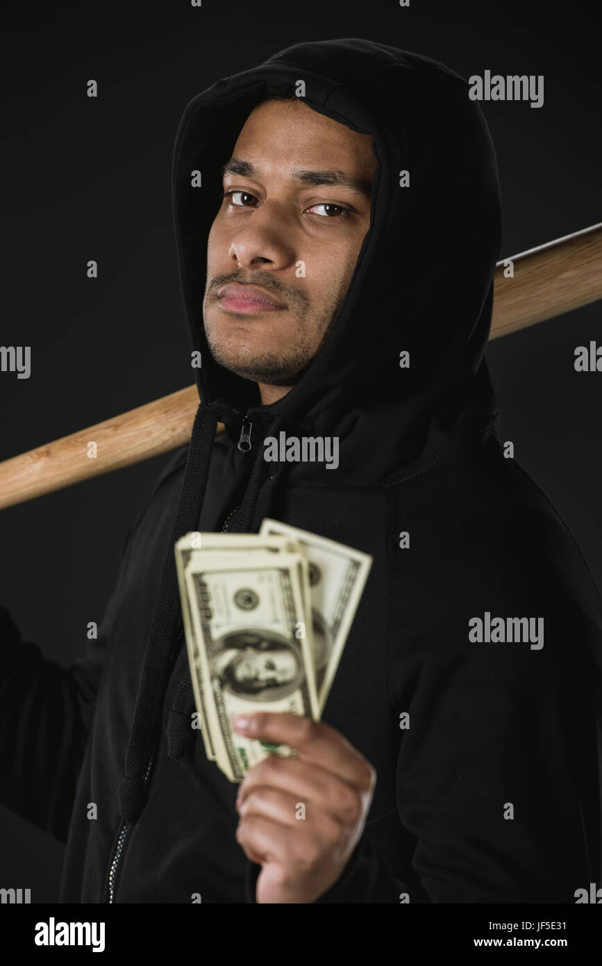 young robber in zoodie with baseball bat and money isolated on black ...