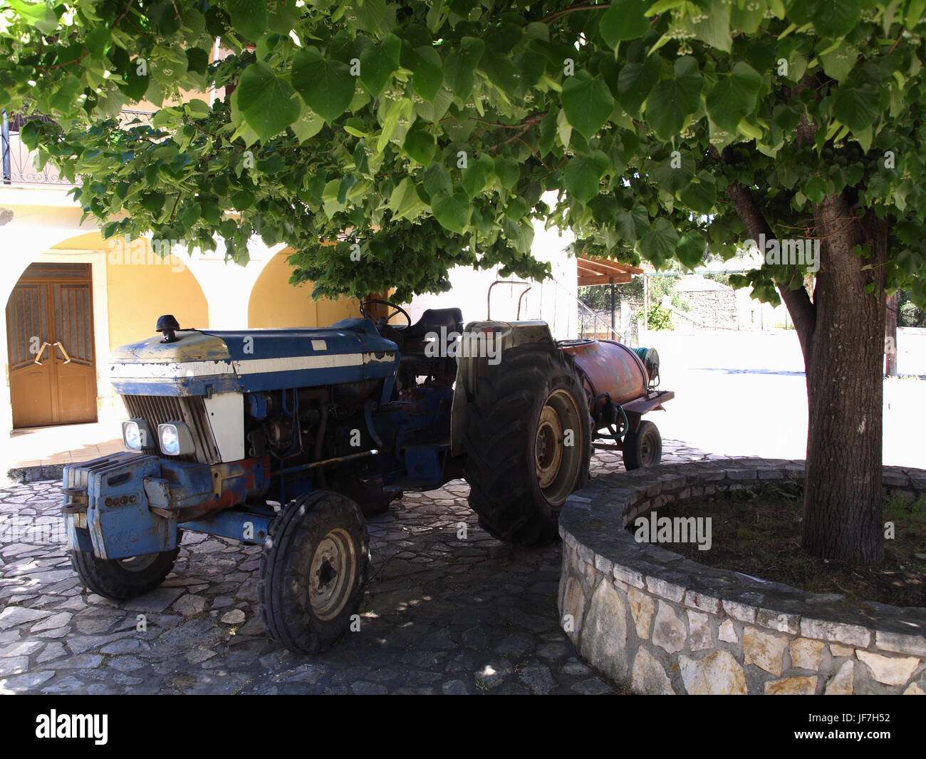 Old tractor parked in shade under tree in village square in Xanthates ...