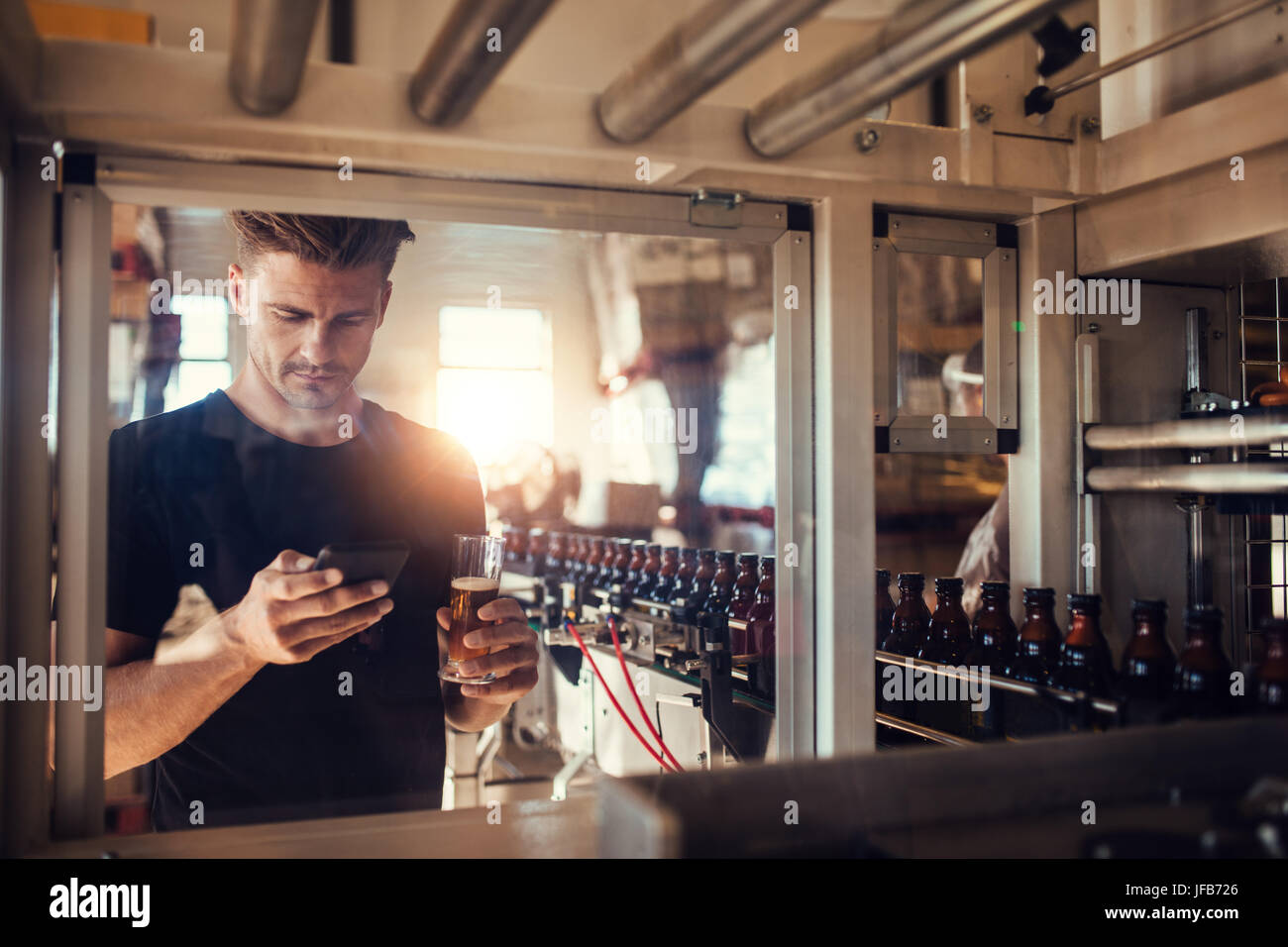 Young man with a glass of beer at brewery plant using mobile phone ...