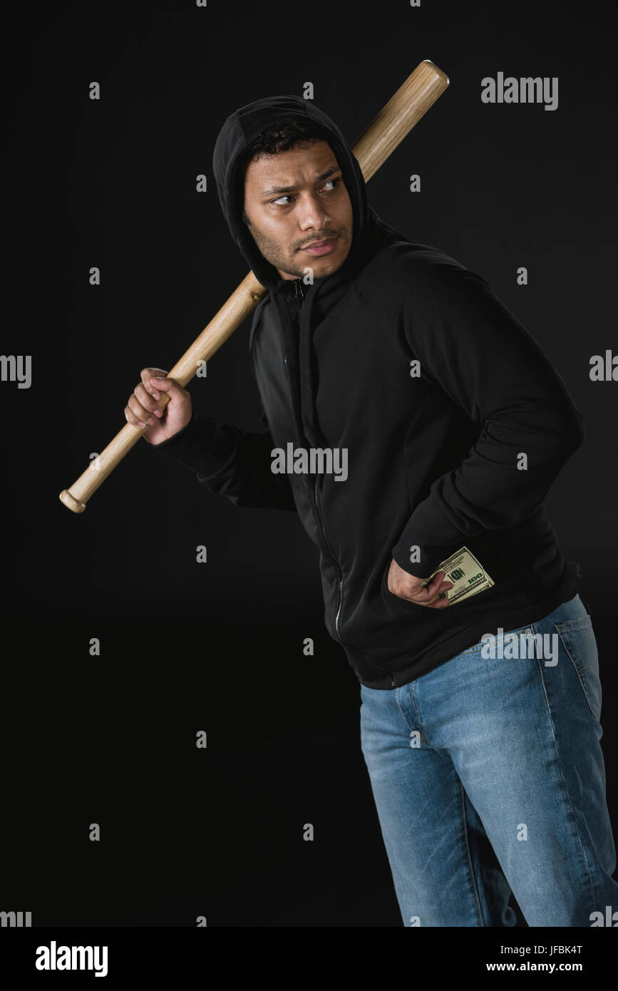 young robber in zoodie with baseball bat and money isolated on black ...